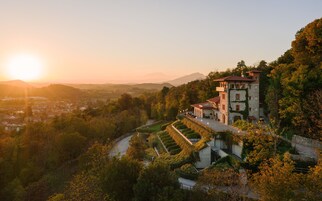 Um edifício histórico com fachada de pedra, jardins em terraços e vista do horizonte durante o pôr do sol.