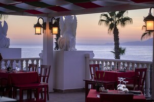 Um restaurante com toalhas e cadeiras vermelhas, um corrimão branco e vista para o mar ao pôr do sol.