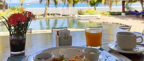 Mesa de café da manhã com ovos mexidos, torradas e uma bebida, com vista para uma praia com palmeiras e uma piscina.