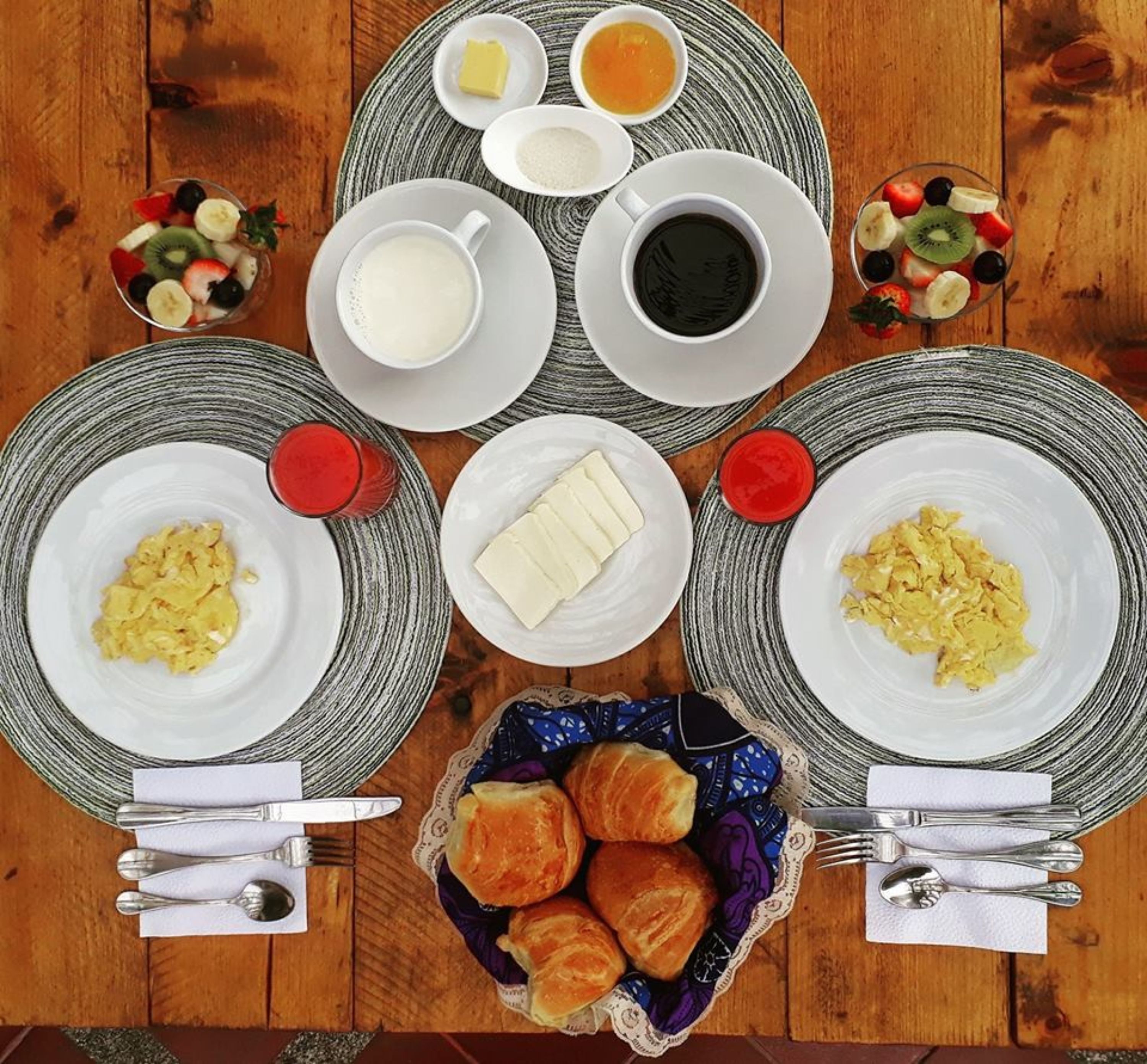 Uma mesa de madeira com ovos mexidos, manteiga, croissants e fruta para o pequeno-almoço.