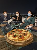 Três pessoas jantando em uma mesa ao ar livre com uma pizza grande à frente.
