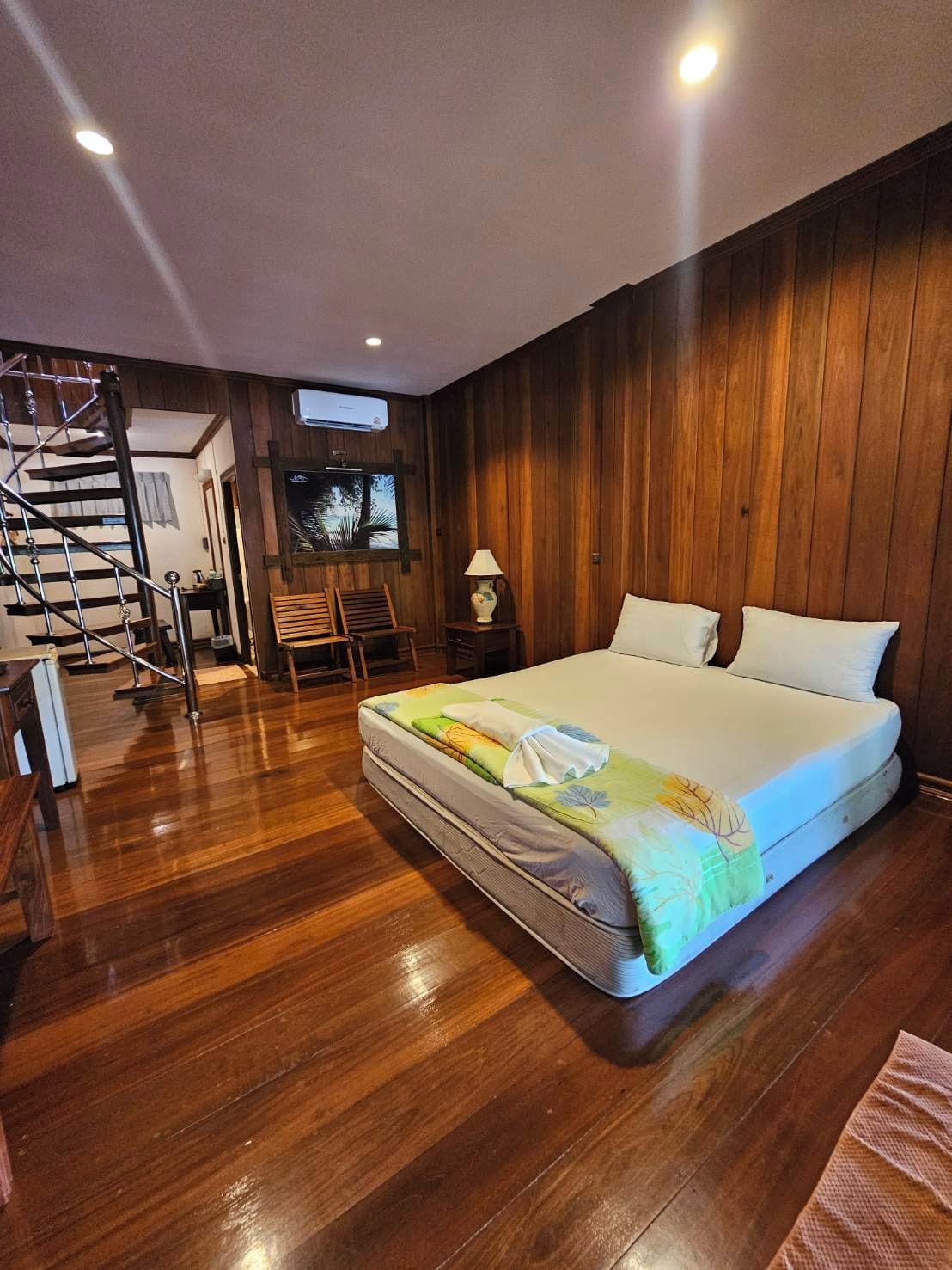 Quarto com piso de madeira, cama com colcha colorida, uma cadeira, luminária e escada.