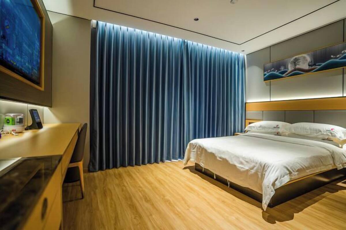 Quarto de hotel moderno com piso de madeira, cama com roupa de cama branca, escrivaninha com computador e uma grande janela com cortinas azuis.