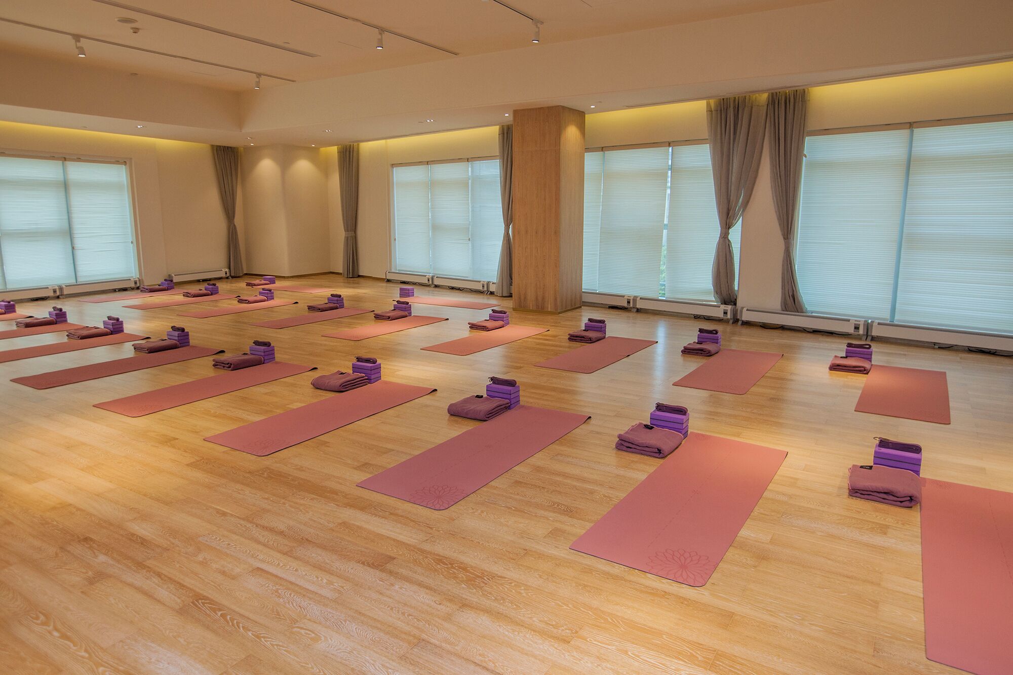 Um quarto espaçoso com fileiras de tapetes e blocos de yoga, grandes janelas com persianas e piso de madeira.