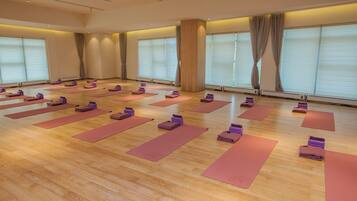 Um quarto espaçoso com fileiras de tapetes e blocos de yoga, grandes janelas com persianas e piso de madeira.