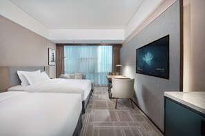 Quarto de hotel com duas camas, uma escrivaninha, uma cadeira, uma televisão e uma janela grande com cortinas.
