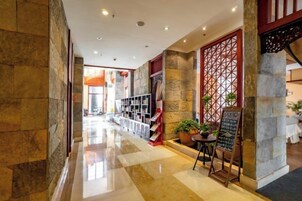 Corredor de hotel com balcão de recepção, área de estar com mesa e cadeiras, e parede decorativa com padrão geométrico.
