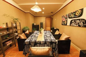 Uma sala de jantar com uma mesa comprida, cadeiras e um lustre. Há prateleiras com plantas e uma porta ao fundo.