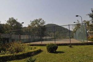 Campo de tênis cercado, céu claro e árvores ao fundo.