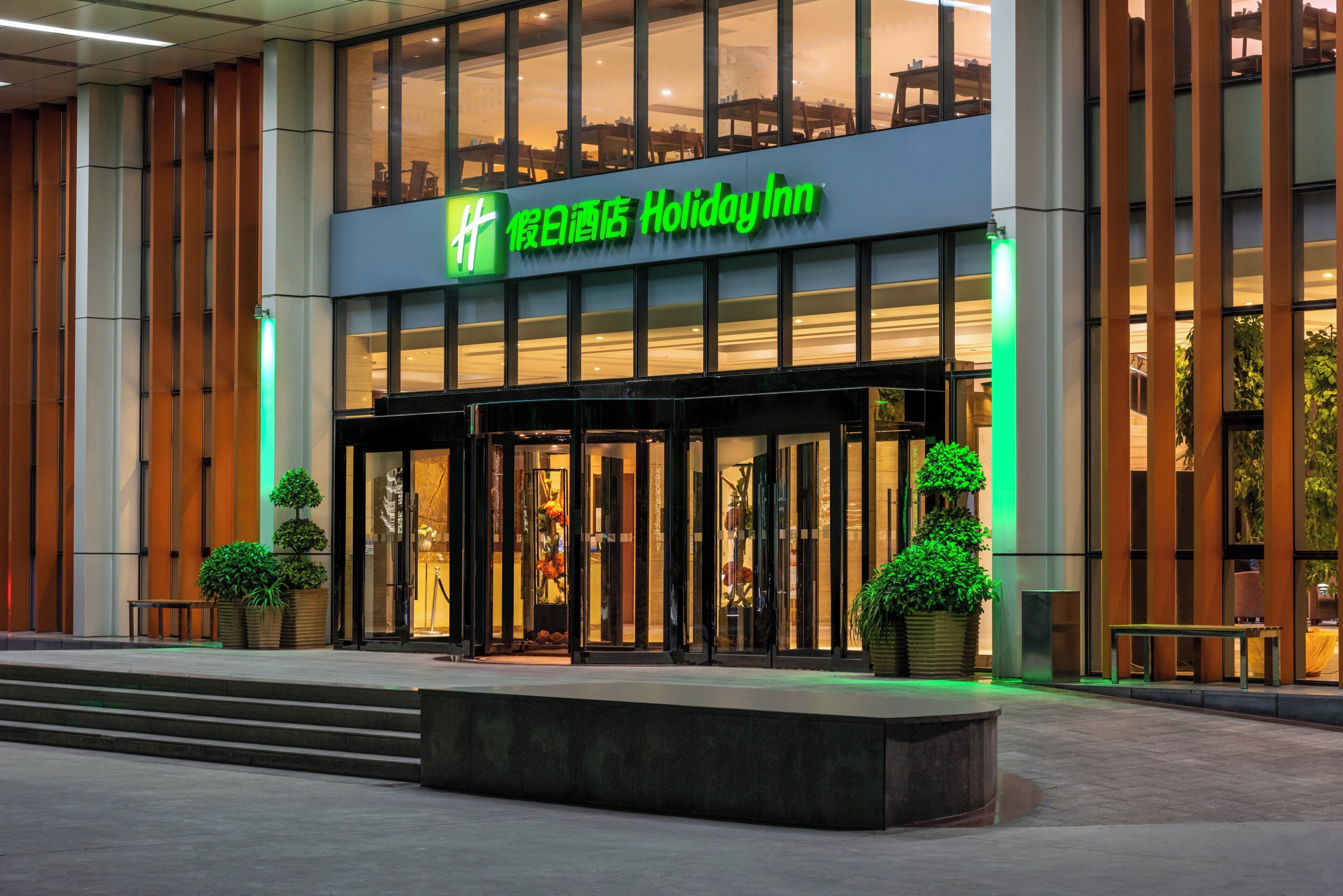 Entrada de um hotel Holiday Inn com fachada de vidro, uma placa verde e uma pequena área externa com vasos de plantas.