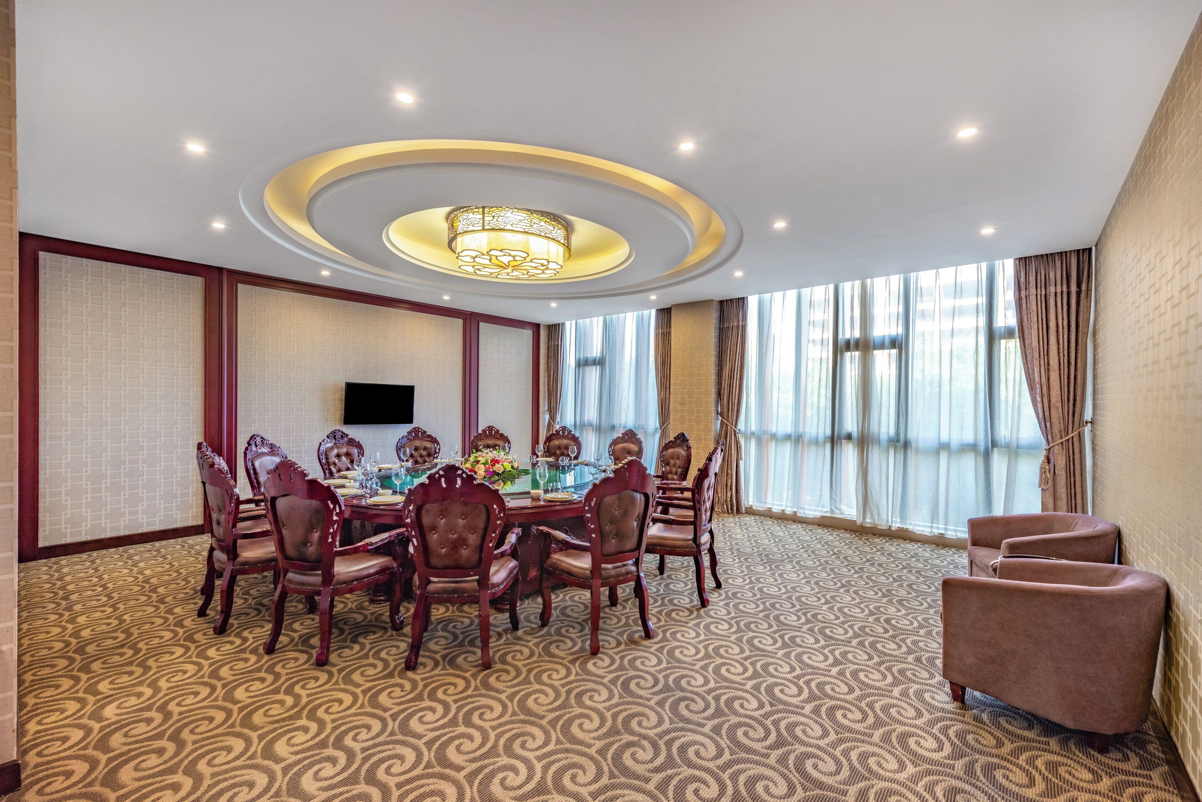 Uma sala de conferências com uma mesa redonda, cadeiras, um candelabro e uma janela grande com cortinas.