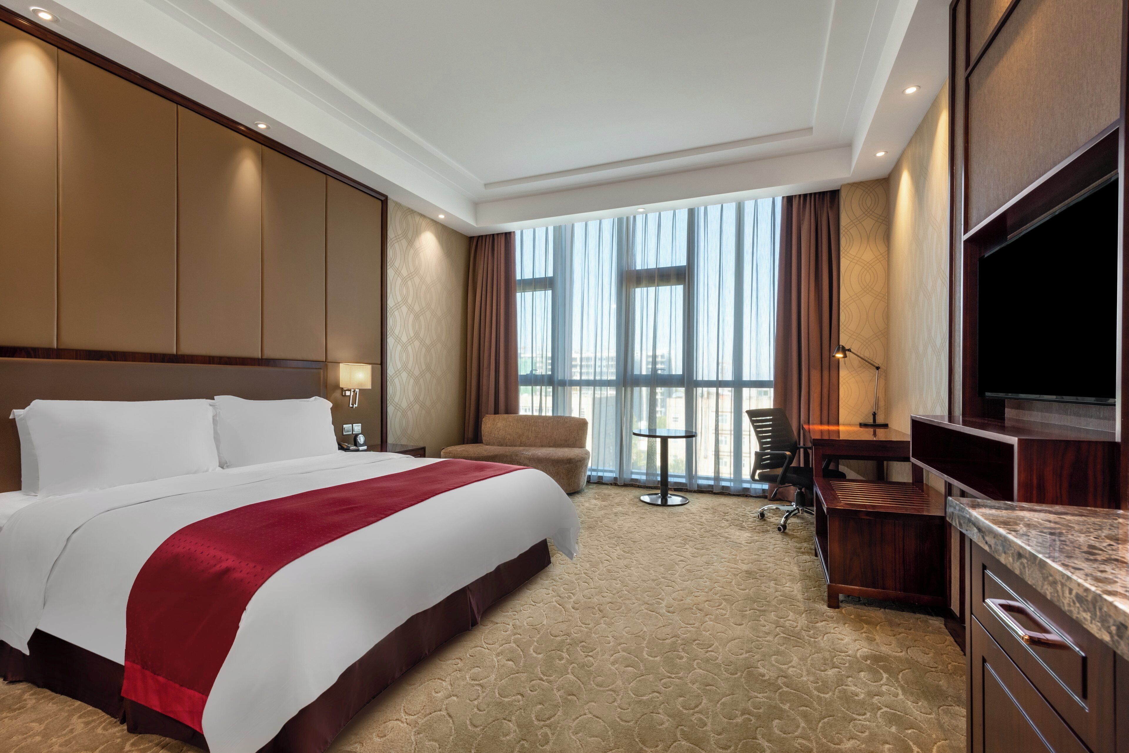 Quarto de hotel com uma cama grande, uma escrivaninha, uma cadeira, uma televisão e vista para a cidade por amplas janelas.