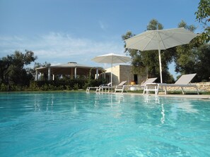 Piscina com espreguiçadeiras e guarda-sóis, cercada por vegetação e um edifício de pedra.