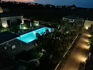 Piscina retangular com espreguiçadeiras e guarda-sóis, cercada por vegetação e um caminho pavimentado.