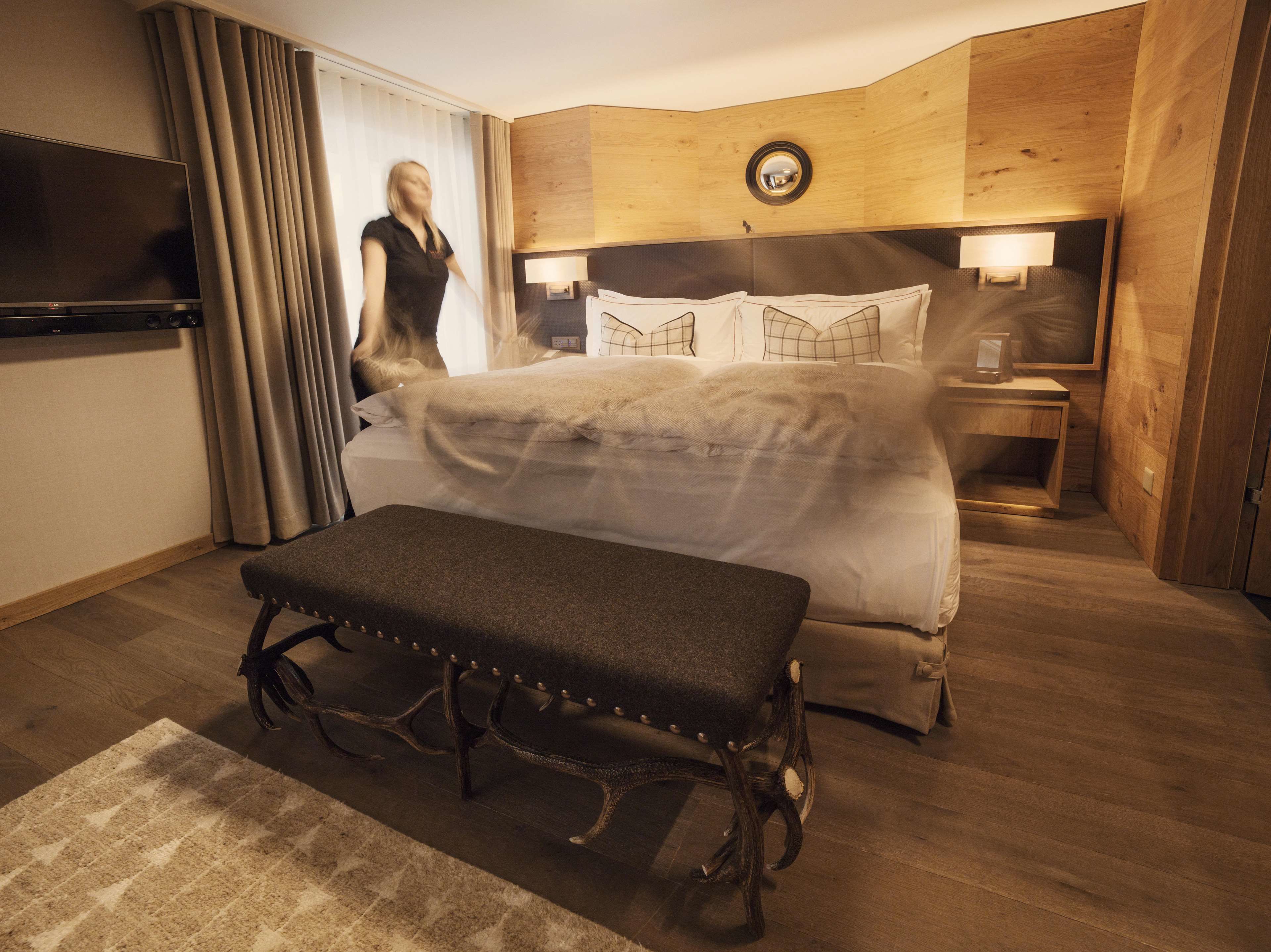 Quarto de hotel com uma cama grande, um banco, uma TV e painéis de madeira.