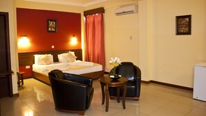 Quarto de hotel com cama, duas cadeiras, uma mesinha, ar condicionado e um quadro na parede.