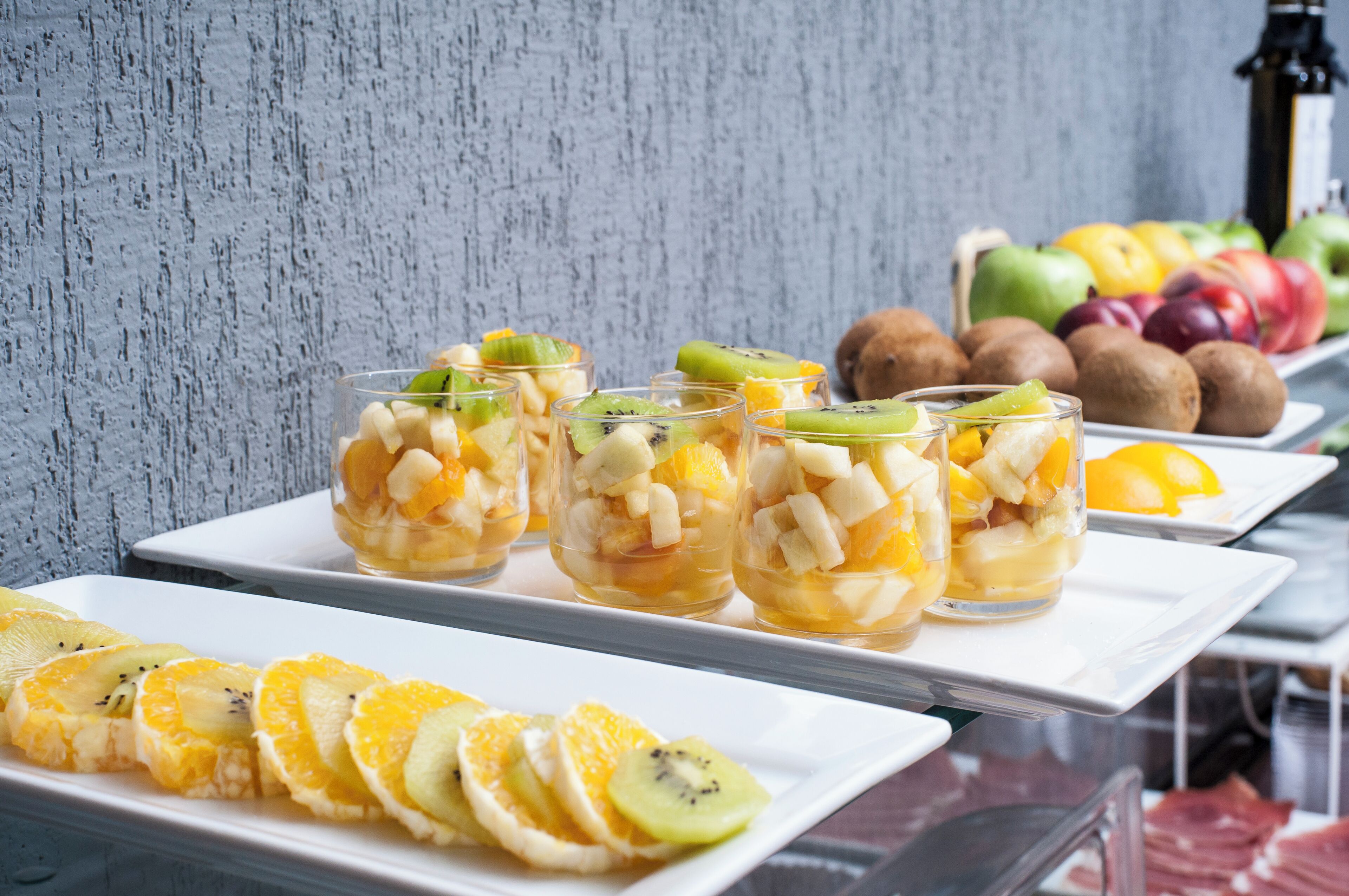 Uma tábua de frutas com laranjas e kiwis fatiados em um prato branco, e uma variedade de frutas inteiras ao fundo.
