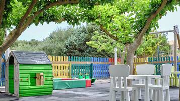 Uma área de recreação infantil com uma casinha verde, cadeiras de plástico branco e um cercado colorido.
