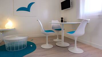 Quarto com parede branca, um mural de baleia azul, uma mesa branca com duas cadeiras, uma pequena estante branca com uma luminária, e uma cadeira branca com almofada azul.