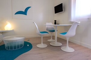 Quarto com parede branca, um mural de baleia azul, uma mesa branca com duas cadeiras, uma pequena estante branca com uma luminária, e uma cadeira branca com almofada azul.