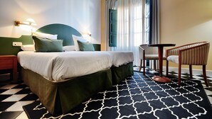 Quarto de hotel com uma cama de casal, uma cadeira, uma mesinha e chão quadriculado.