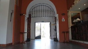 Um saguão de hotel com uma grande entrada em arco, uma recepção e uma escadaria que leva aos andares superiores.