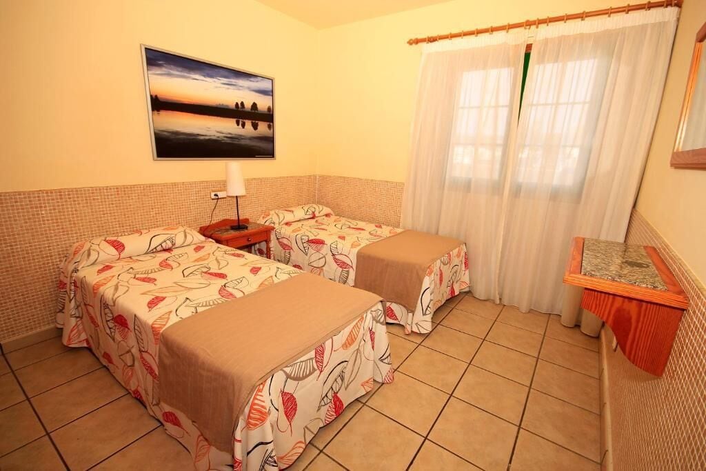 Quarto de hotel com duas camas, mesa de cabeceira, quadro de um pôr do sol e janela com cortinas leves.