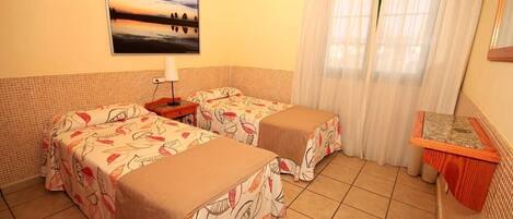 Quarto de hotel com duas camas, mesa de cabeceira, quadro de um pôr do sol e janela com cortinas leves.