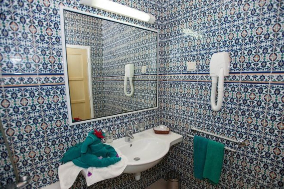 Casa de banho com azulejos estampados, pia, espelho, porta-toalhas e secador de cabelo.