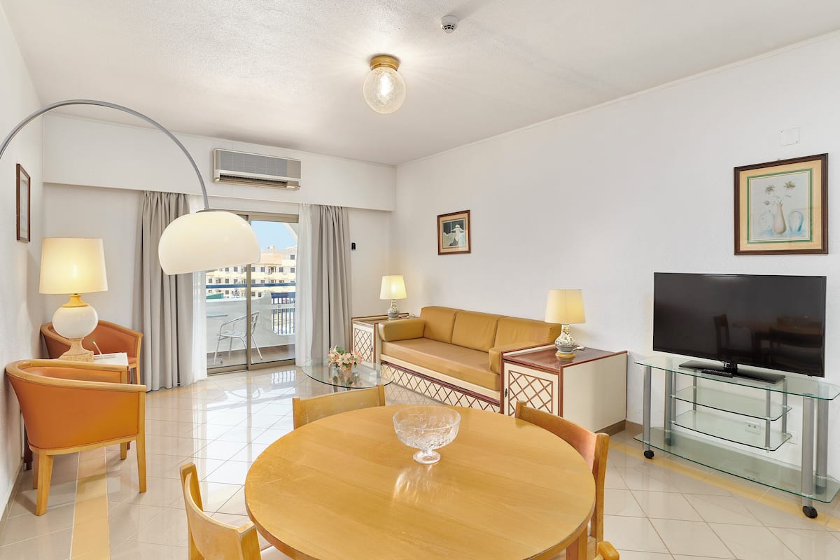 Quarto de hotel com mesa de jantar redonda, duas cadeiras, um sofá, uma TV sobre um suporte de vidro e uma varanda com vista.