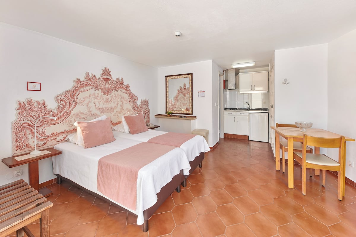 Quarto com uma cama grande, mesas de cabeceira, um banquinho de madeira, uma mesa de jantar com cadeiras e uma cozinha compacta ao fundo.