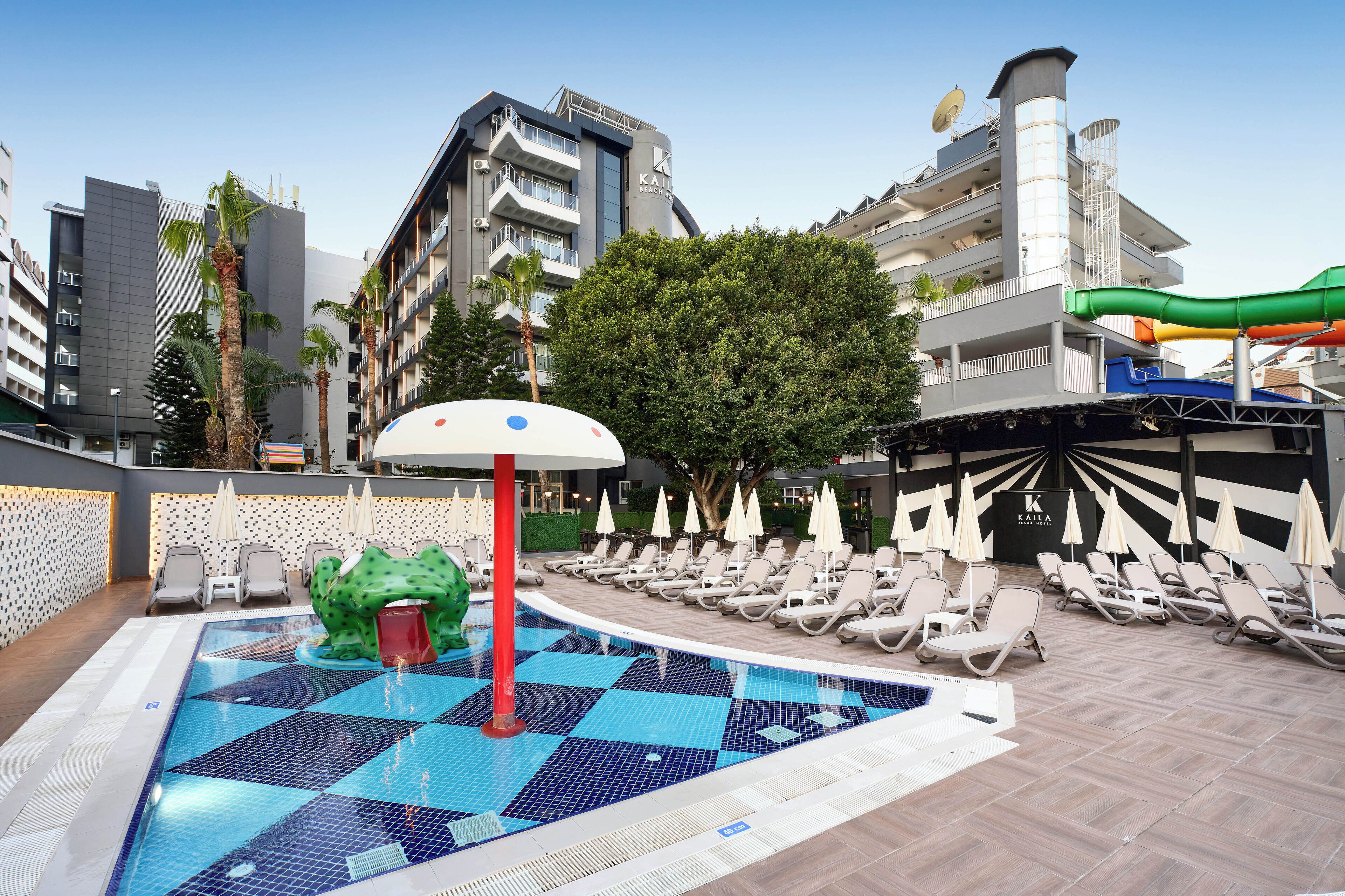 Uma área de piscina no terraço com espreguiçadeiras, um tobogã e um edifício com varandas ao fundo.