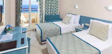 Quarto de hotel com duas camas, uma mesa com um computador e um controle remoto, uma cadeira e vista para o oceano através de uma porta de vidro.