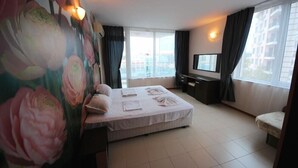 Quarto de hotel com cama, televisão, escrivaninha e uma janela grande.