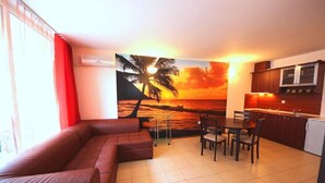 Uma sala de estar com sofá, mesa de jantar com cadeiras, área de cozinha e uma grande pintura de um pôr do sol sobre o oceano.