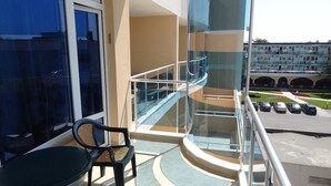 Uma varanda com guarda-corpo de vidro, uma cadeira e vista para um estacionamento e edifícios ao fundo.
