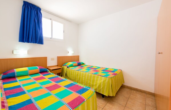 Quarto com duas camas, cada uma com colcha colorida, uma janela com cortinas azuis e uma mesa de cabeceira com abajur.