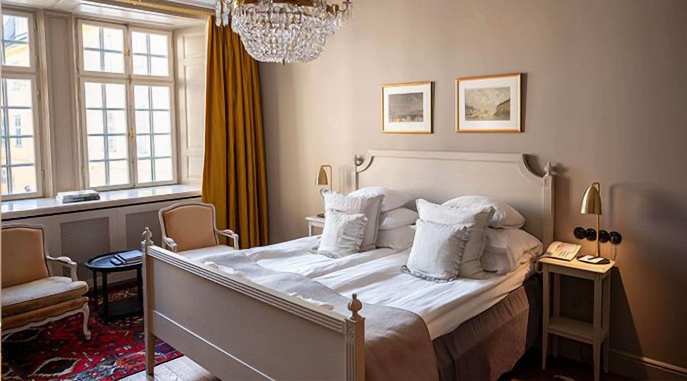 Quarto com cama, mesas de cabeceira, uma cadeira, janela com cortinas e duas imagens emolduradas na parede.