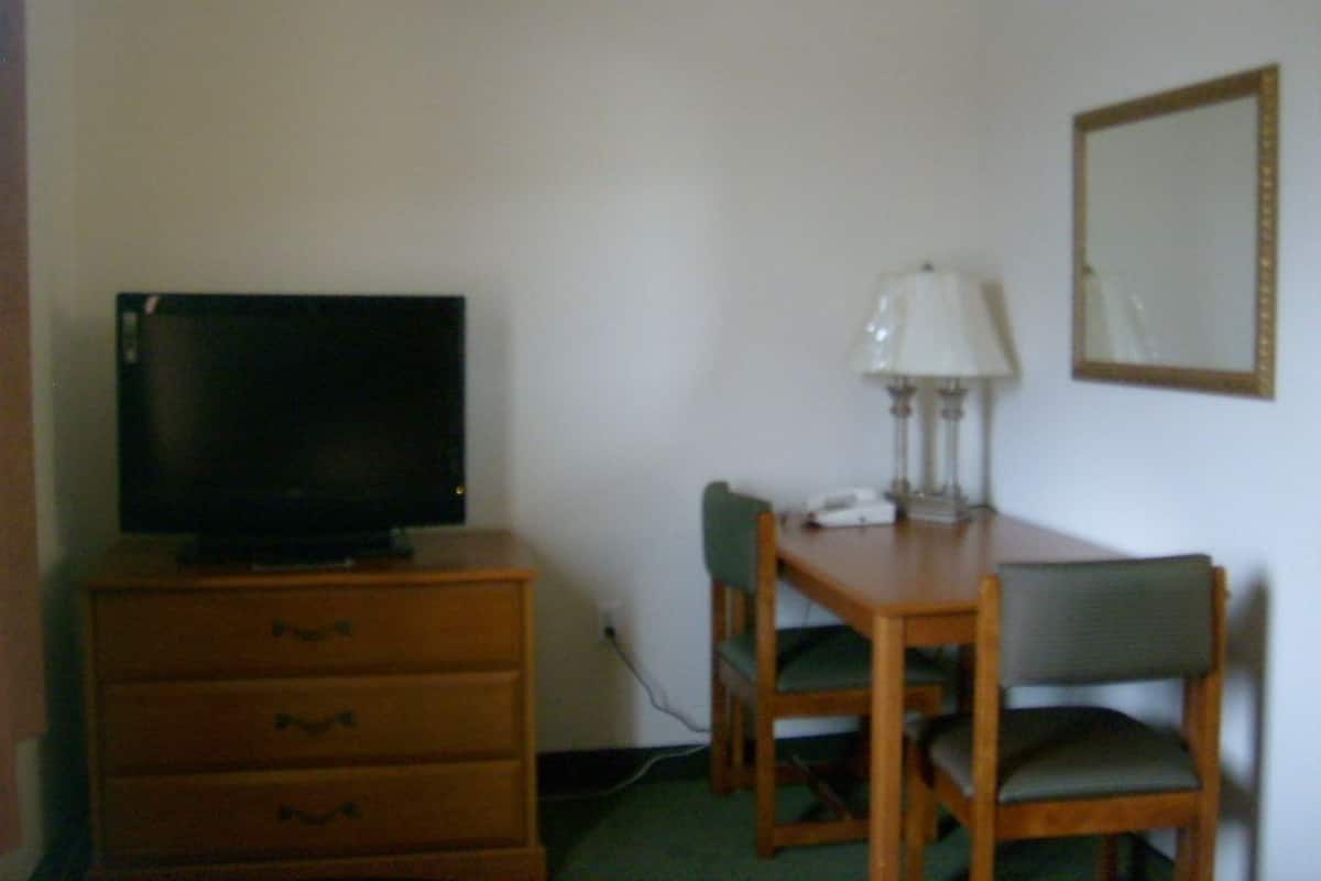 Quarto com televisão sobre uma cômoda de madeira, uma mesa com abajur e uma cadeira.