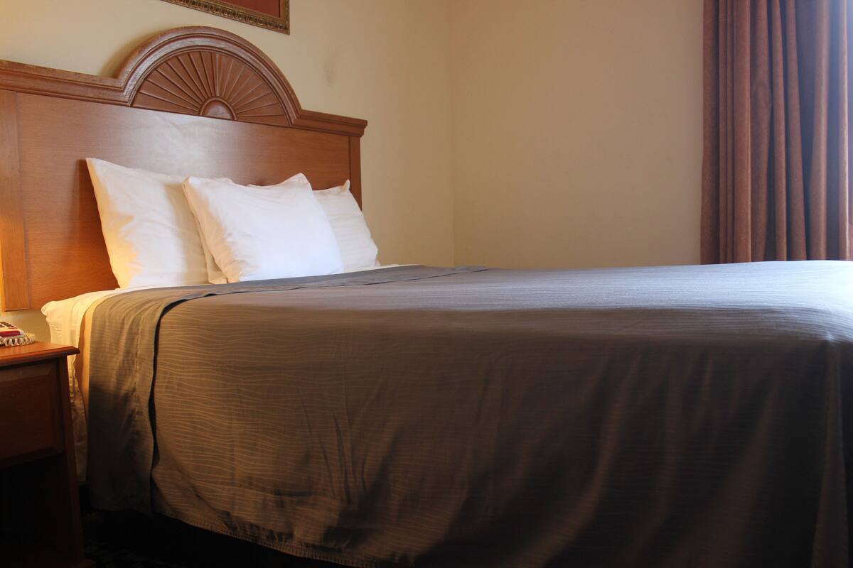 Uma cama bem arrumada com cabeceira de madeira, mesa de cabeceira com abajur e uma janela com cortinas.