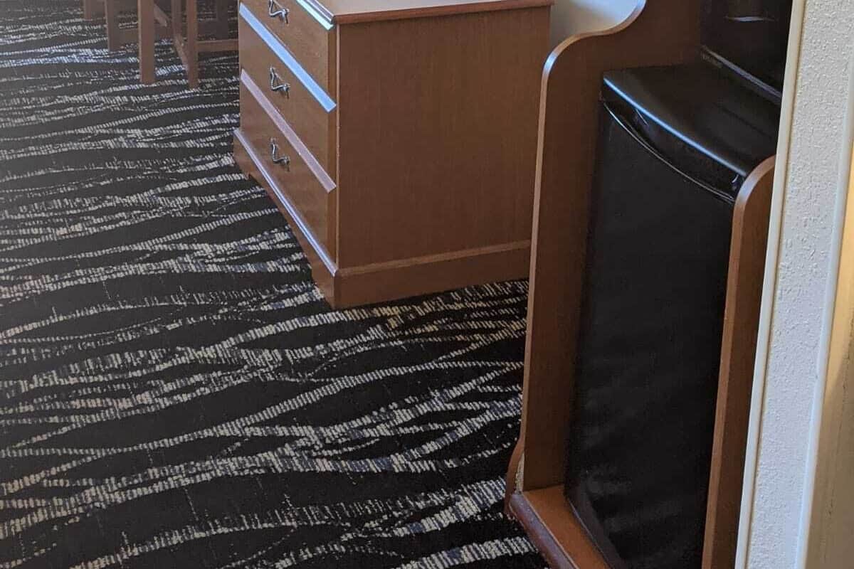 Quarto de hotel com escrivaninha de madeira, televisão fixada na parede, luminária e lixeira.