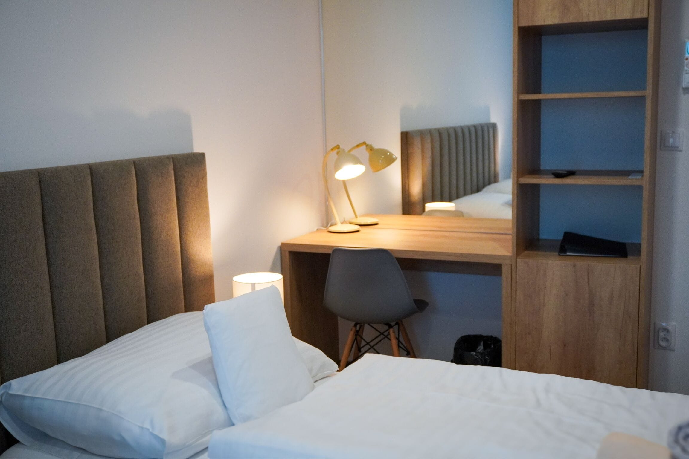 Quarto de hotel com uma cama, escrivaninha, cadeira, abajur e estante de livros.