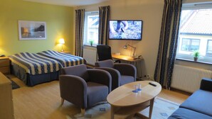 Quarto de hotel com uma cama, duas poltronas, uma escrivaninha, uma televisão e uma janela com cortinas.