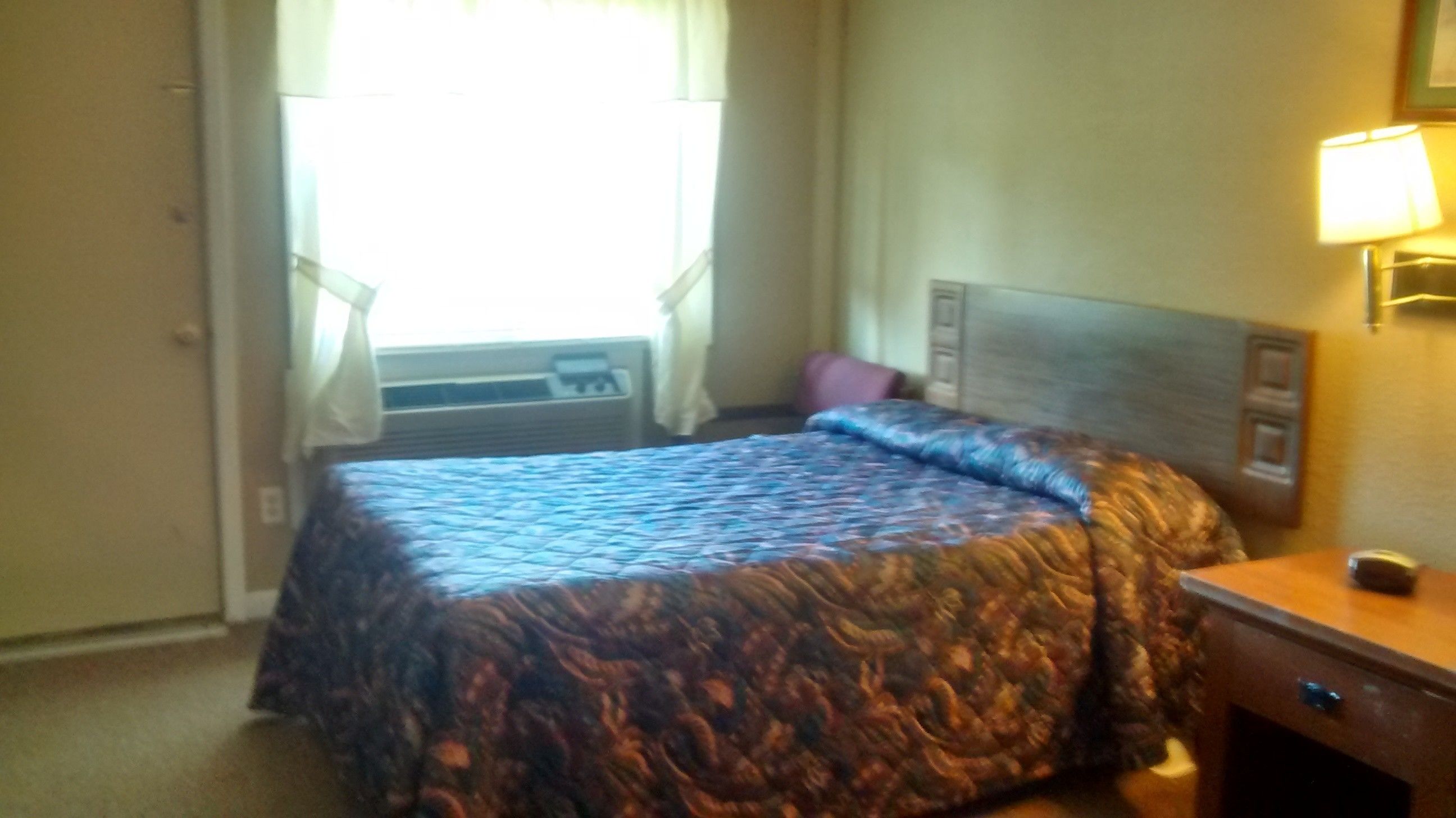 Quarto de hotel com cama, escrivaninha, cadeira, luminária e janela com cortinas.