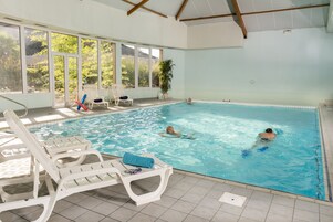 Piscina interna com água azul cristalina, rodeada por cadeiras de lounge brancas e uma grande janela com vista para o verde lá fora.