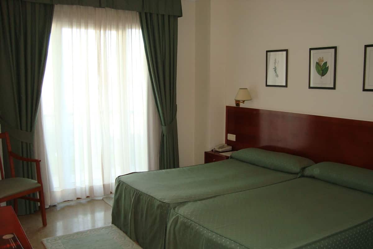 Quarto de hotel com duas camas, uma mesa de cabeceira de madeira com abajur, uma cadeira e uma janela com cortinas.