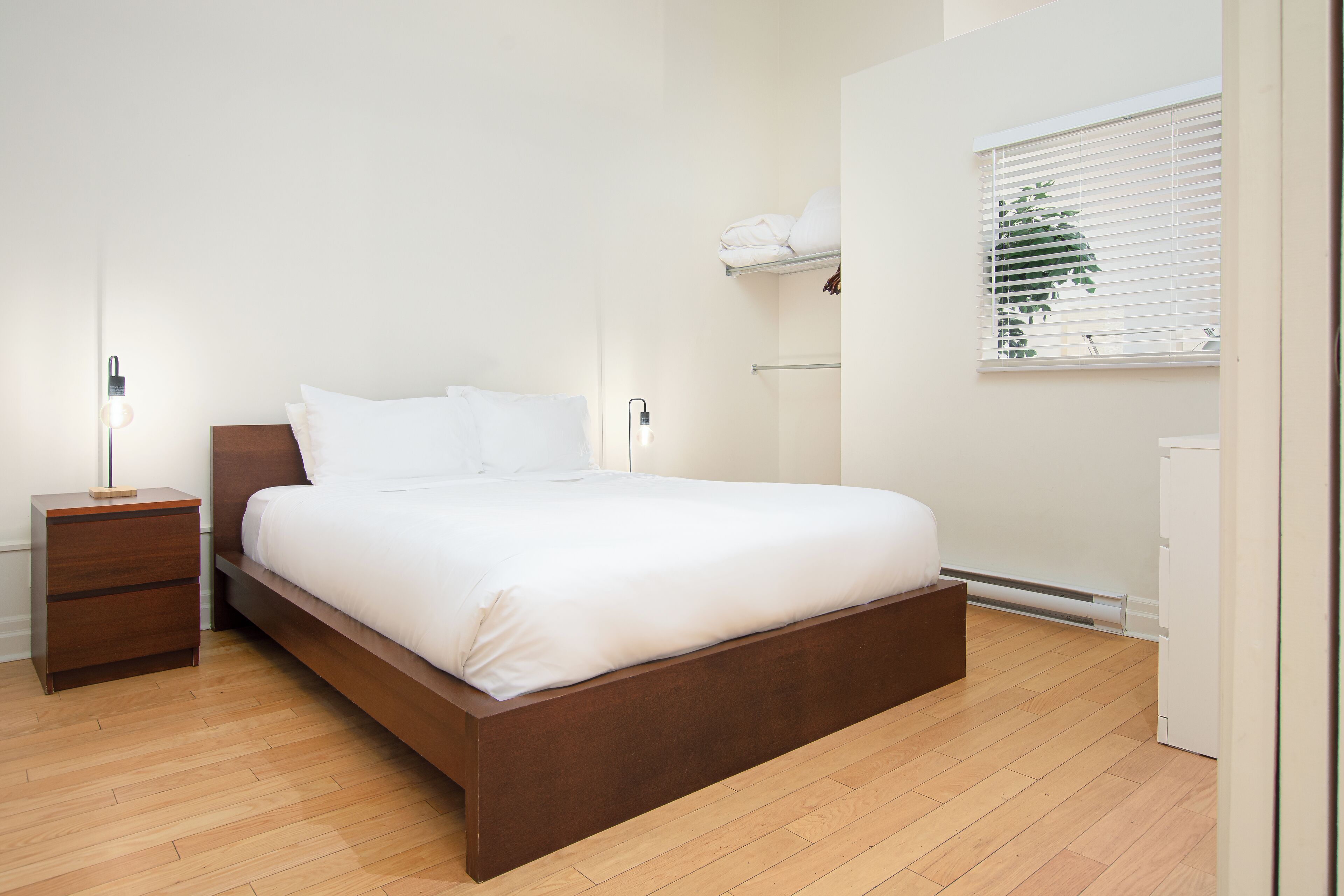 Quarto com cama de madeira, mesas de cabeceira, janela com persianas e radiador.
