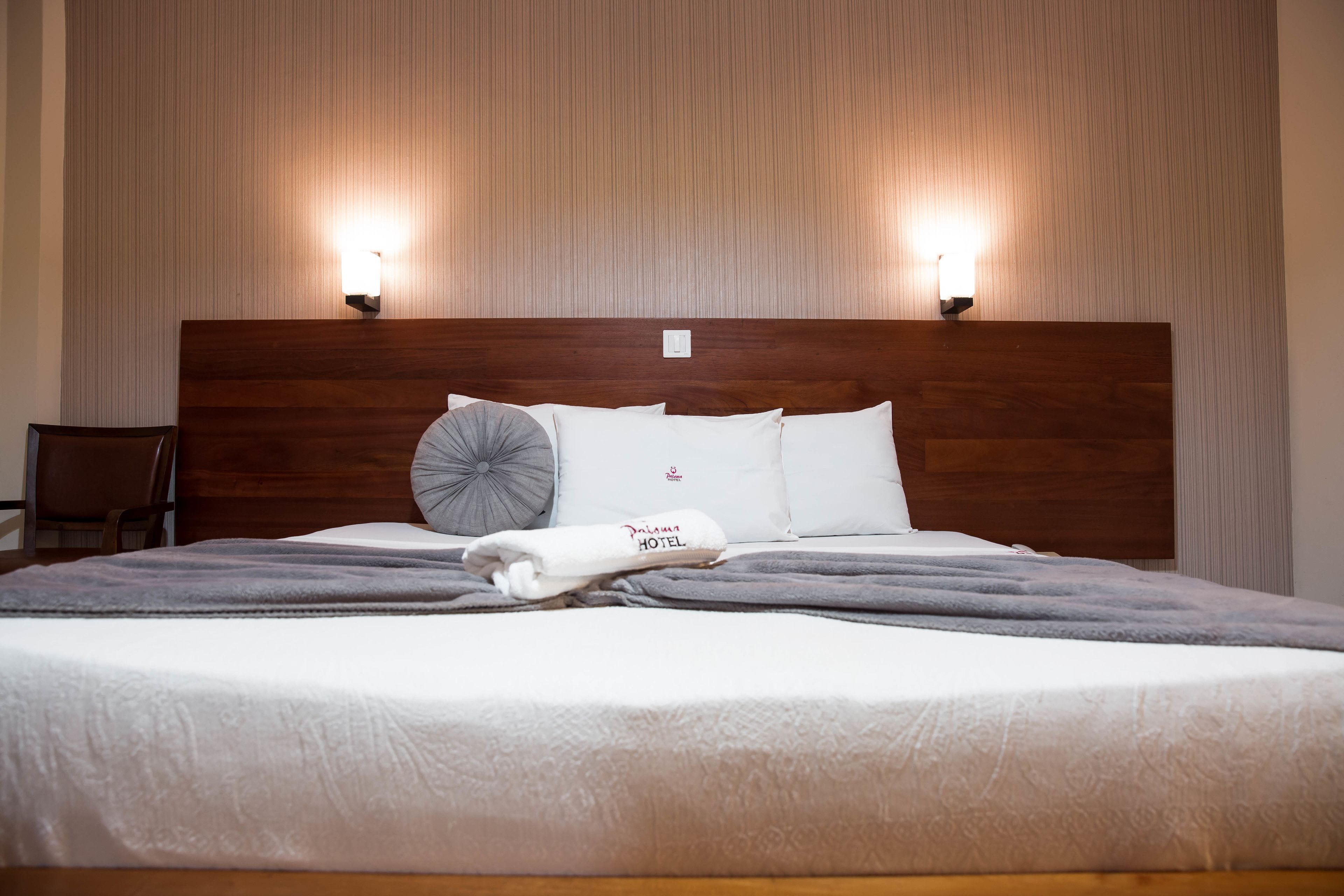 Cama bem arrumada com cabeceira de madeira, duas luminárias fixadas na parede e uma toalha dobrada com a inscrição "Dalmia Hotel".