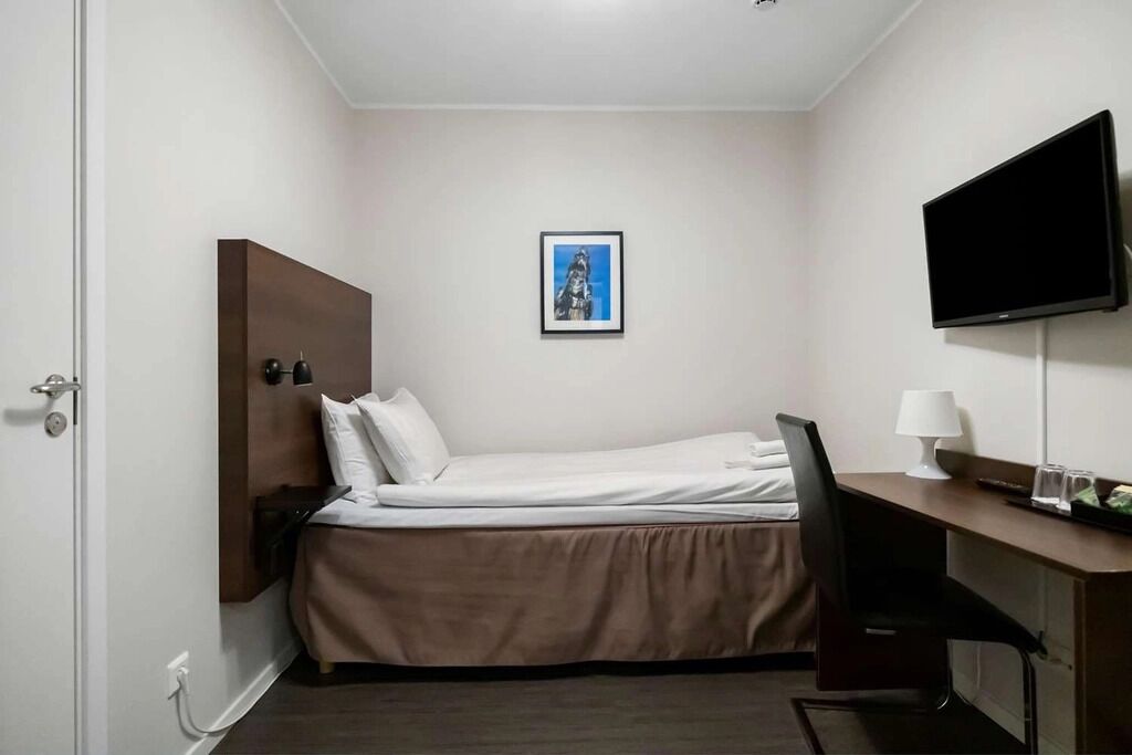 Quarto de hotel pequeno e bem arrumado com uma cama de solteiro, uma escrivaninha com cadeira, uma TV de tela plana e um quadro na parede.