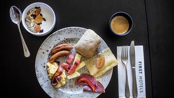 Prato de café da manhã com ovos, linguiça, queijo e frutas, acompanhado de uma tigela de cereal e uma xícara de café sobre uma mesa escura.